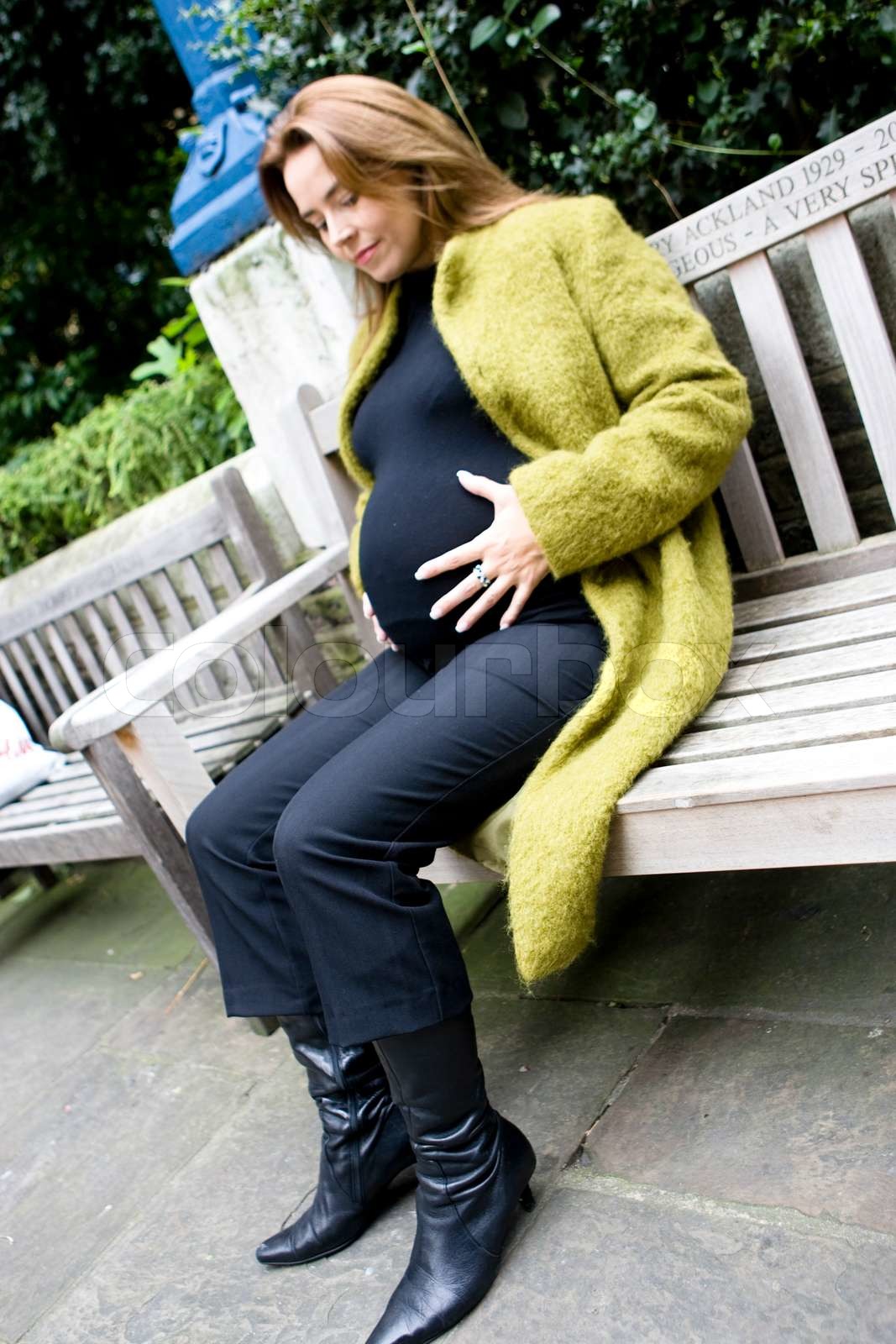 A Pregnant Woman Holds Her Stomach While Sitting On Bench In A Park