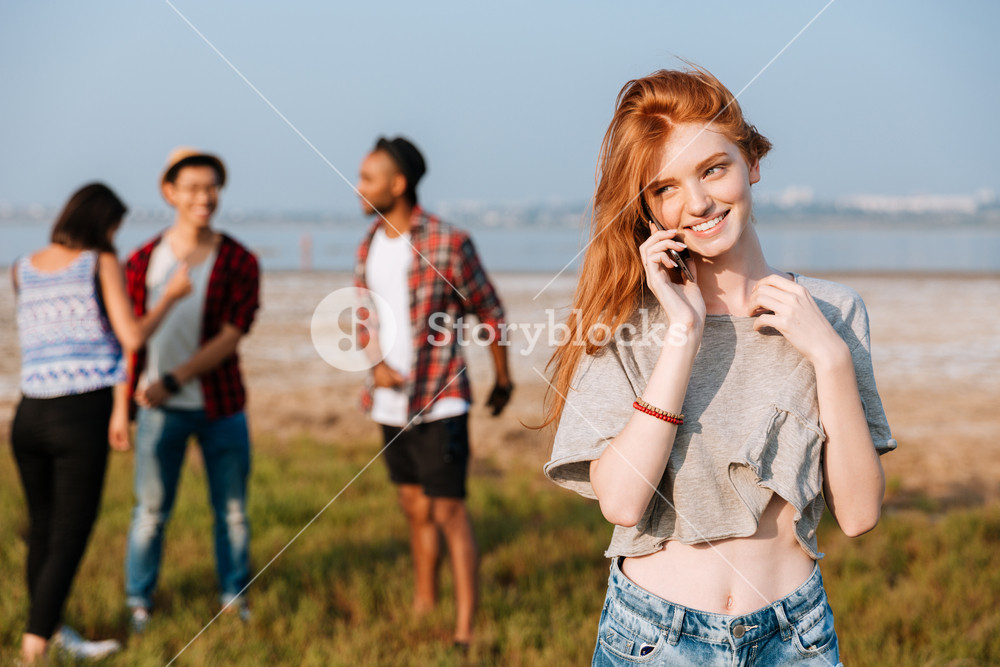 Cheerful Redhead Young Woman Talking On Cell Phone Standing Near Her